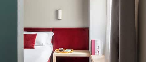 Quarto de hotel com cama, mesa de cabeceira, pequena mesa com bandeja, luminária na parede e janela com cortinas.