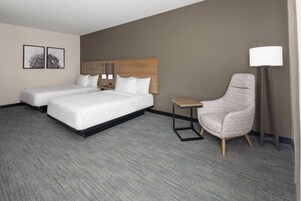 Quarto de hotel com duas camas, uma cadeira, uma mesinha e um abajur.