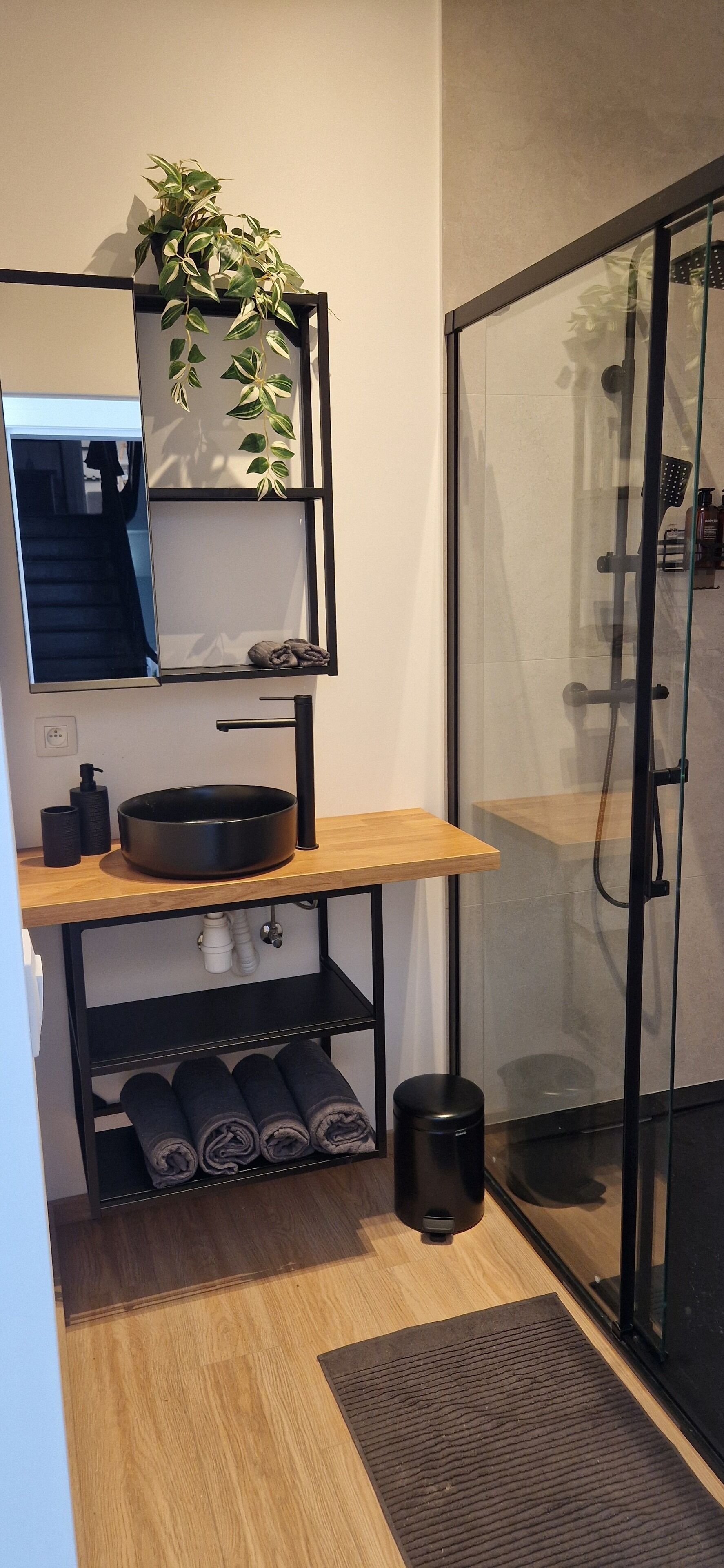 Casa de banho compacta com toucador de madeira, lavatório preto, espelho, planta num vaso, cabine de duche em vidro e um contentor de lixo preto.