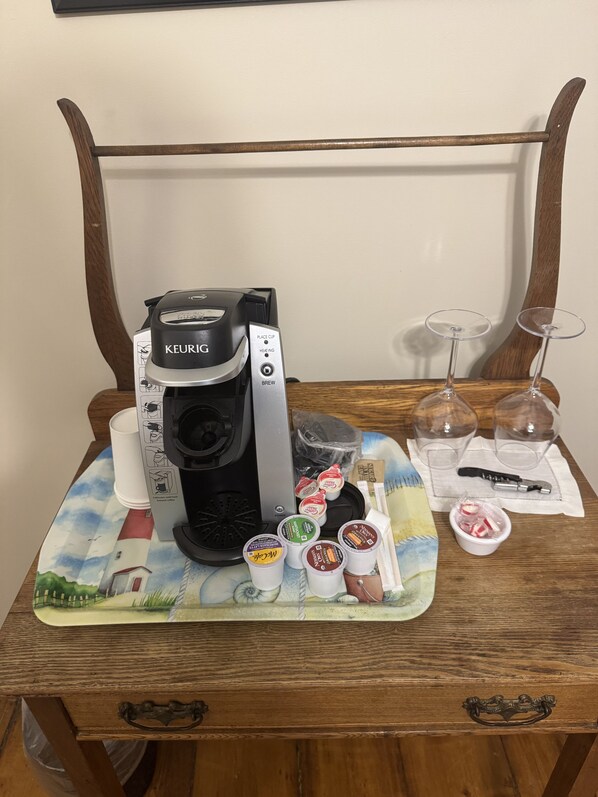 Uma cafeteira sobre uma mesa de madeira com um apoio para prato, dois copos de vinho, uma faca e vários sachês de café.