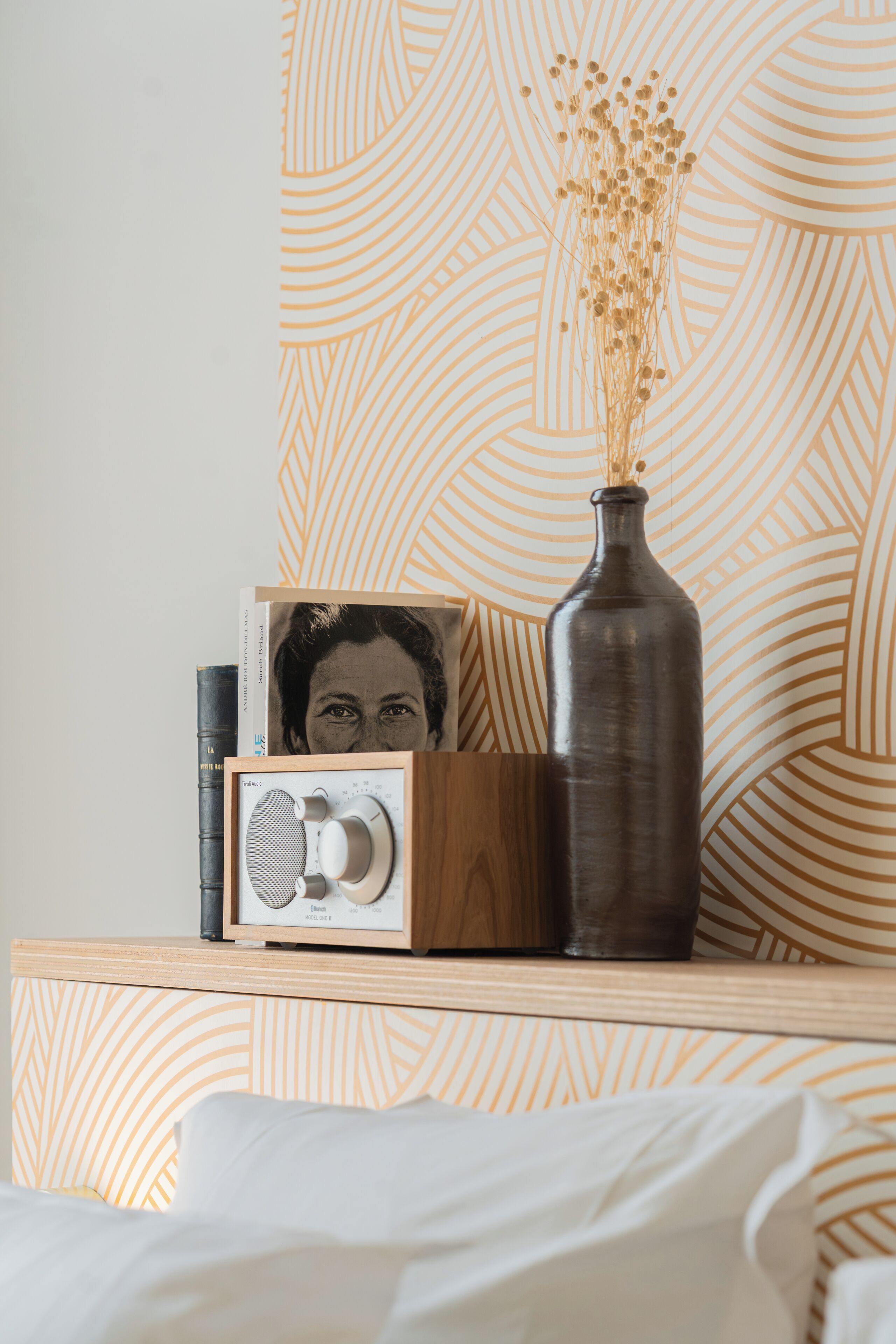 Uma mesinha de cabeceira com um rádio, livros, um vaso com plantas secas e uma parede estampada.