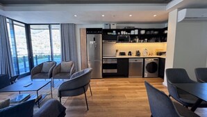 Uma sala de estar moderna com um sofá, cadeiras e uma mesa de centro. Uma cozinha com eletrodomésticos e uma área de jantar com cadeiras são visíveis ao fundo.