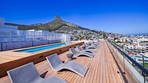 Área de piscina no terraço com espreguiçadeiras e vista para montanhas e paisagem urbana.