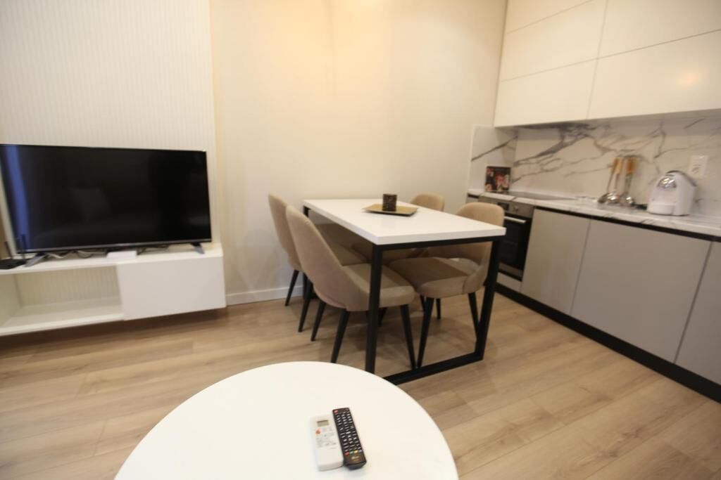 Uma cozinha moderna com uma bancada de mármore, uma área de jantar com uma mesa e cadeiras brancas, e uma TV fixada em um armário branco.