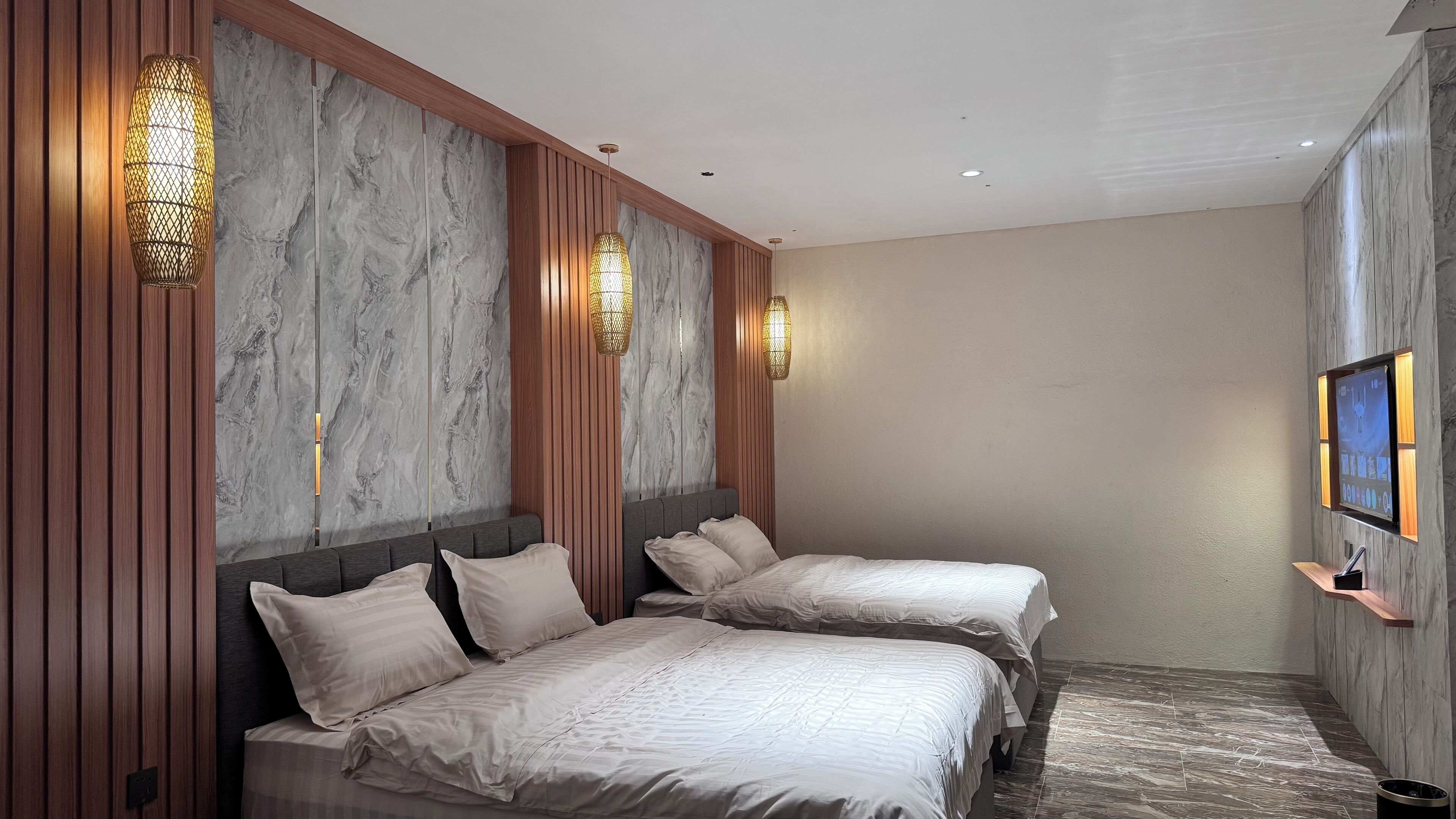 Quarto de hotel com duas camas, painéis de madeira, parede de mármore e uma televisão fixada.