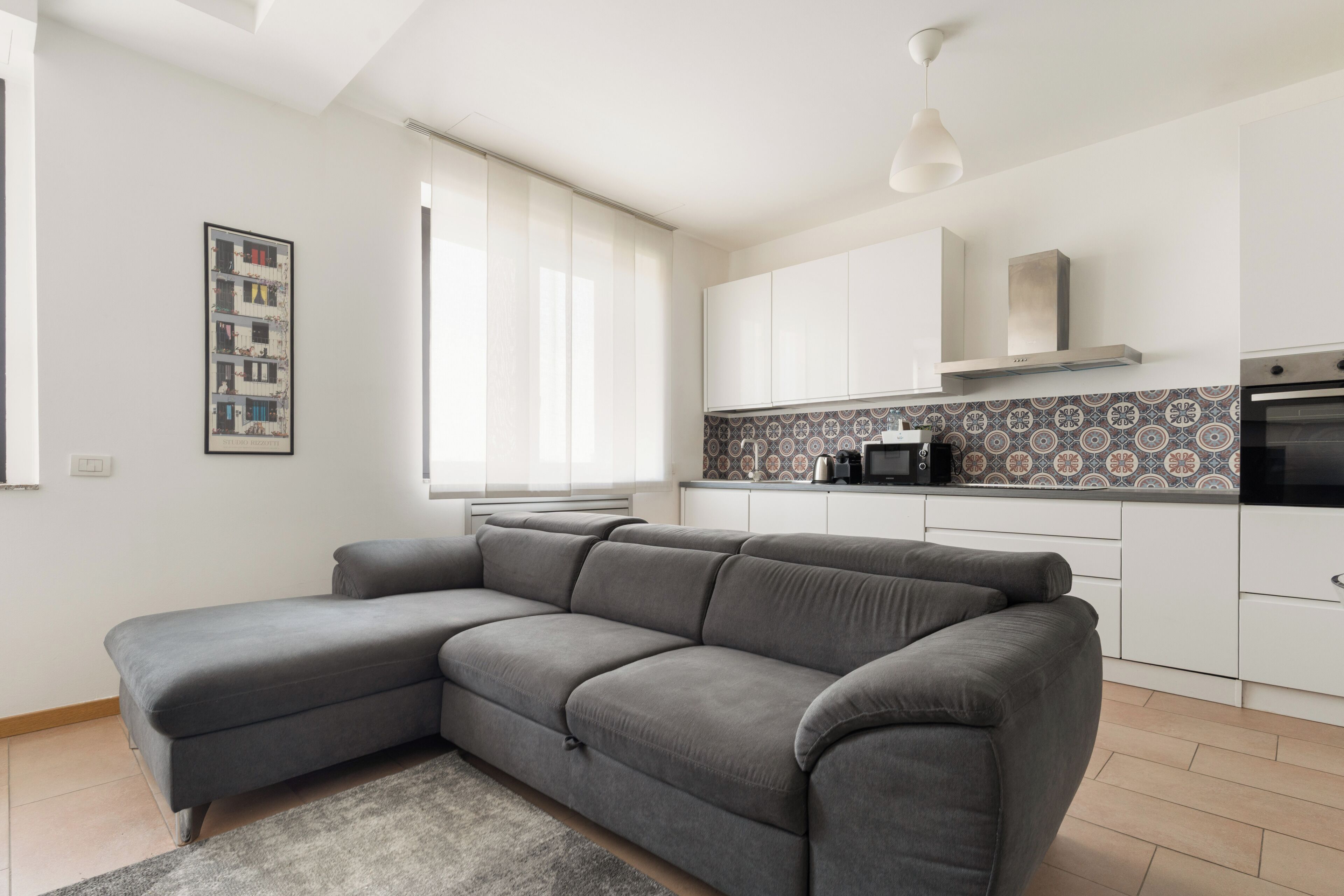 Uma sala de estar moderna com um sofá seccional cinza, uma cozinha com armários brancos e painel de fundo trabalhado, e um espaço de jantar com mesa e cadeiras.