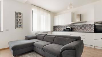 Uma sala de estar moderna com um sofá seccional cinza, uma cozinha com armários brancos e painel de fundo trabalhado, e um espaço de jantar com mesa e cadeiras.