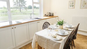 Uma sala de jantar com mesa posta para quatro pessoas, uma janela com vista para um campo gramado e um balcão de cozinha com chaleira e torradeira.