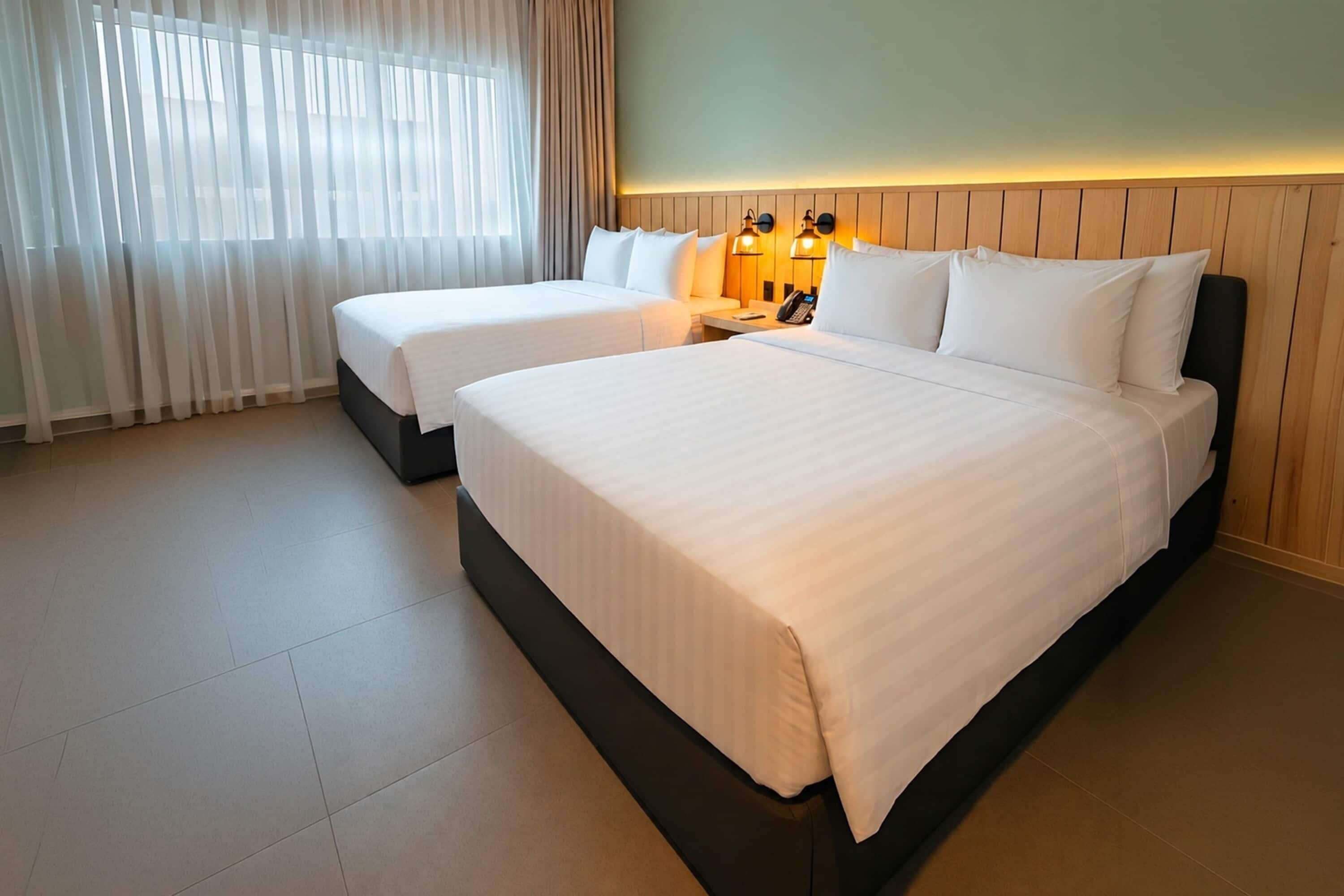 Quarto de hotel com duas camas, cabeceira de madeira, telefone e janela com cortinas leves.
