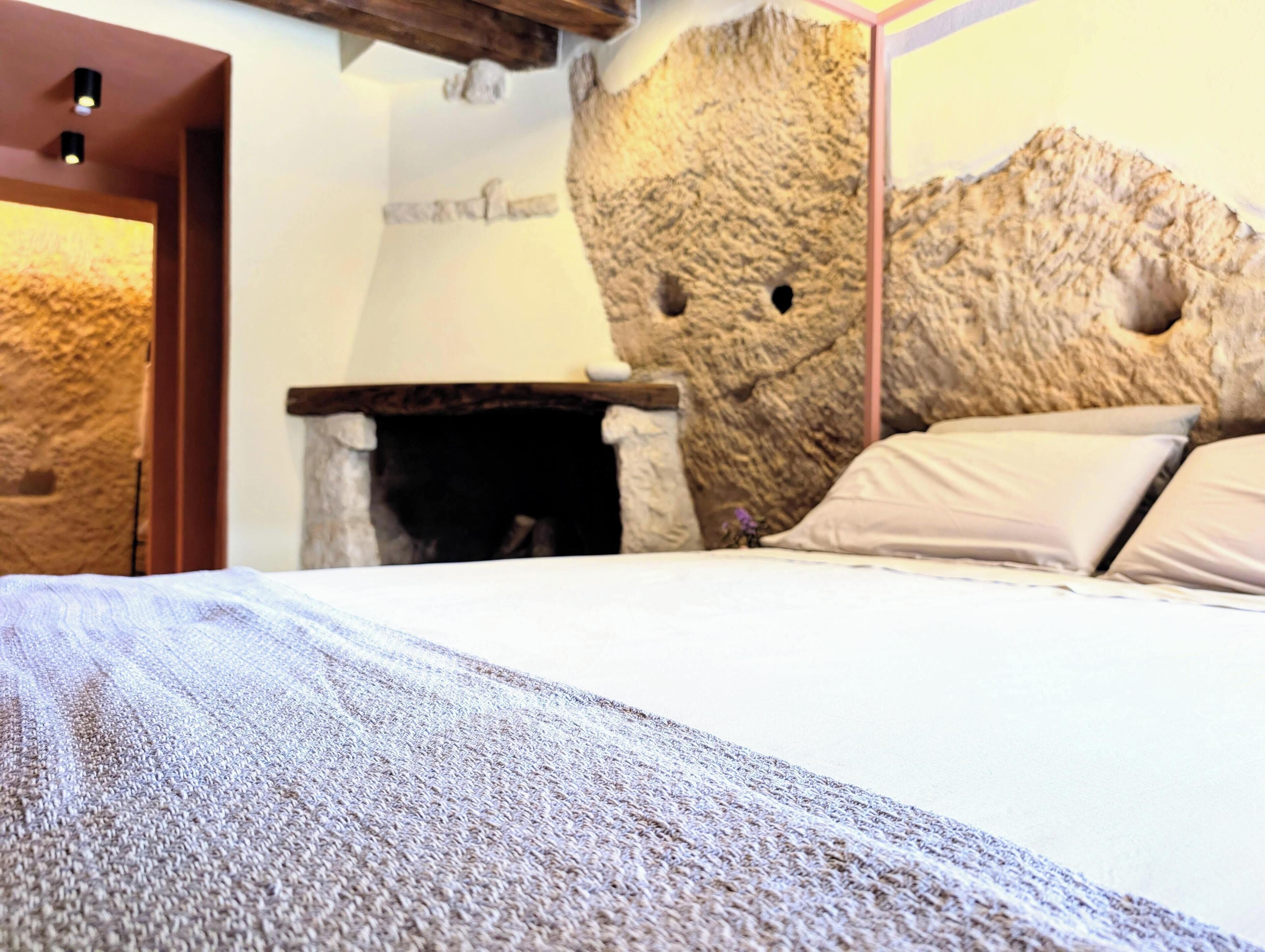 Quarto com uma grande cabeceira de pedra texturizada, cama com roupa de cama branca e uma pequena mesa de cabeceira de madeira com uma luminária.