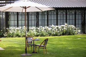Uma área de estar ao ar livre com mesa e cadeiras sob um grande guarda-sol, rodeada por um gramado bem cuidado e flores brancas.