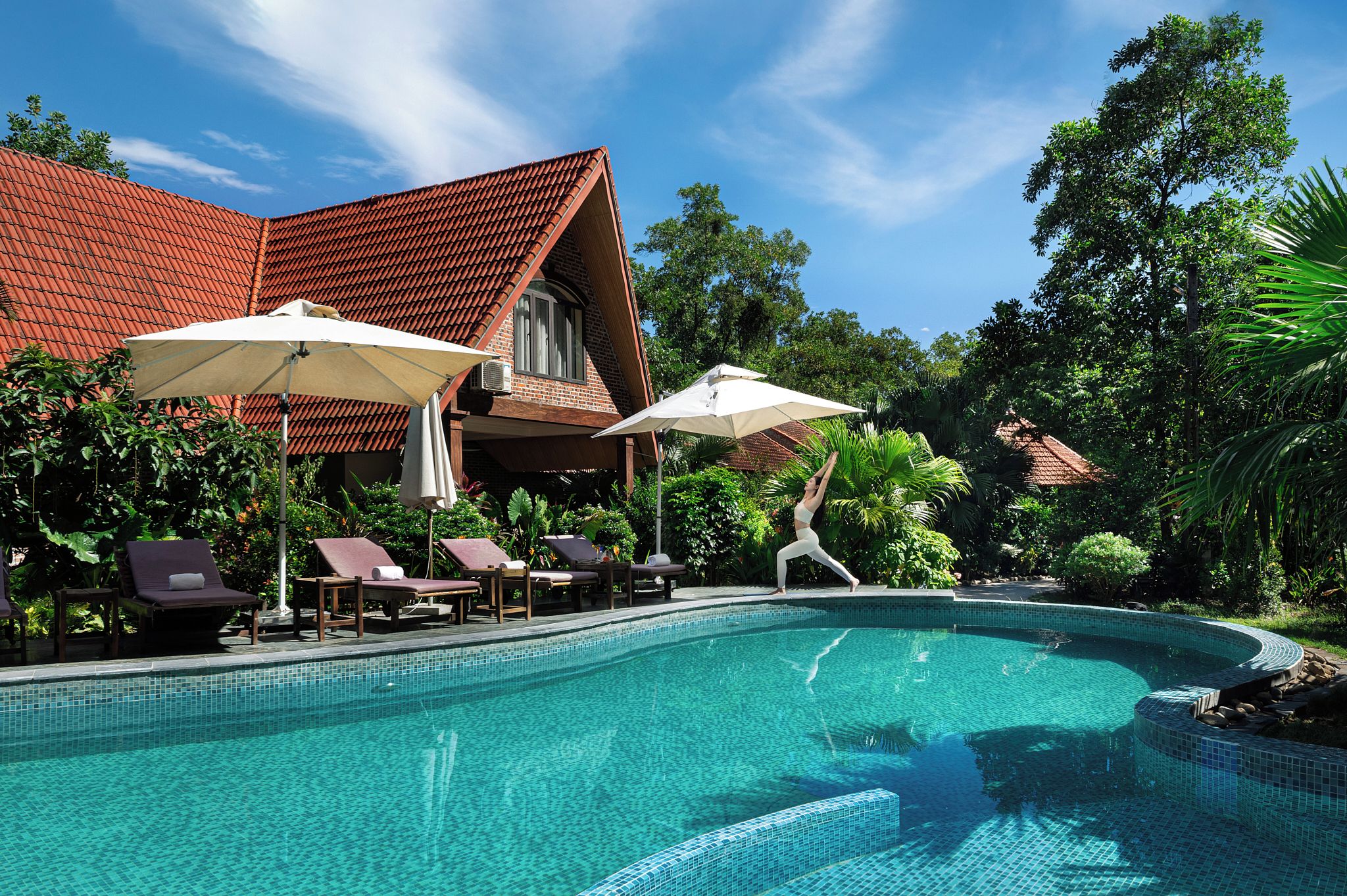 Piscina com espreguiçadeiras e guarda-sóis, cercada por vegetação exuberante e um edifício com telhado em azulejos.