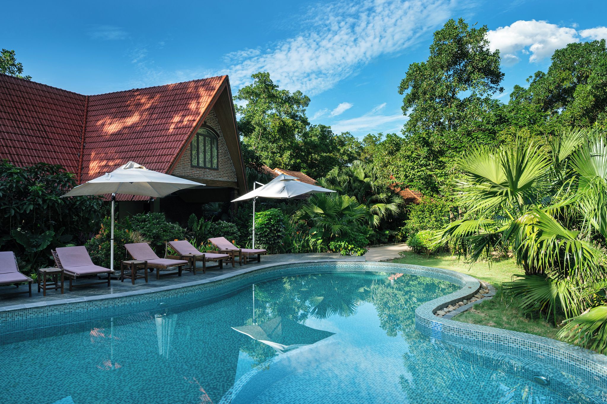Piscina com espreguiçadeiras e guarda-sóis, cercada por vegetação exuberante e um edifício com telhado vermelho.
