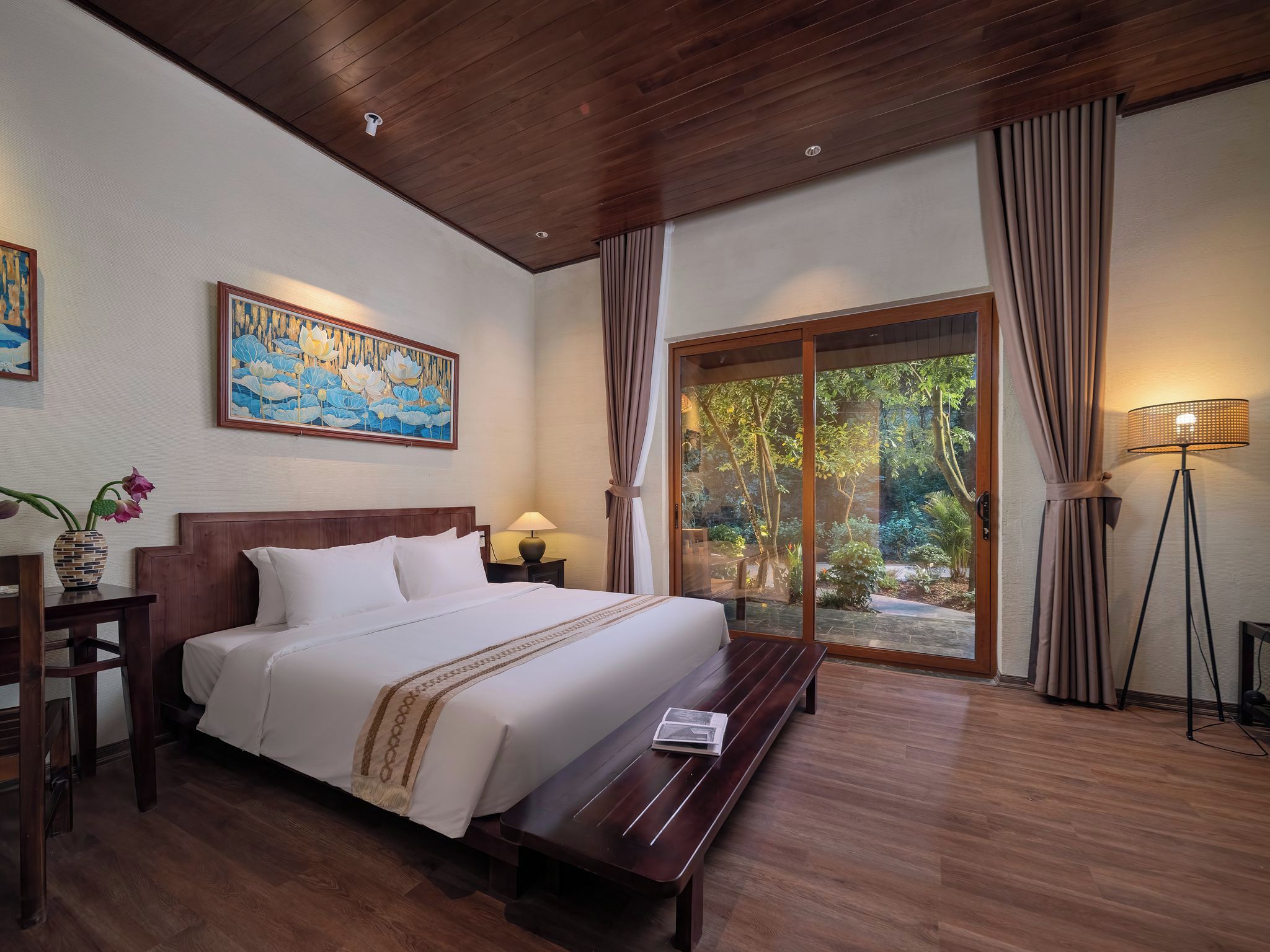 Quarto de hotel com teto de madeira, uma cama grande com roupa de cama branca, um banco, uma mesa de cabeceira com abajur, um quadro na parede e vista para o verde através de uma porta de correr.