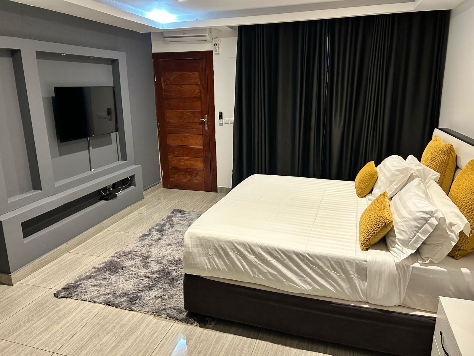 Quarto moderno com uma cama grande, uma TV de tela plana na parede, uma porta de madeira e uma cortina preta.