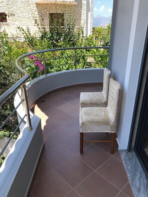 Varanda curva com cadeira estofada, grade de metal e um edifício de pedra ao fundo.
