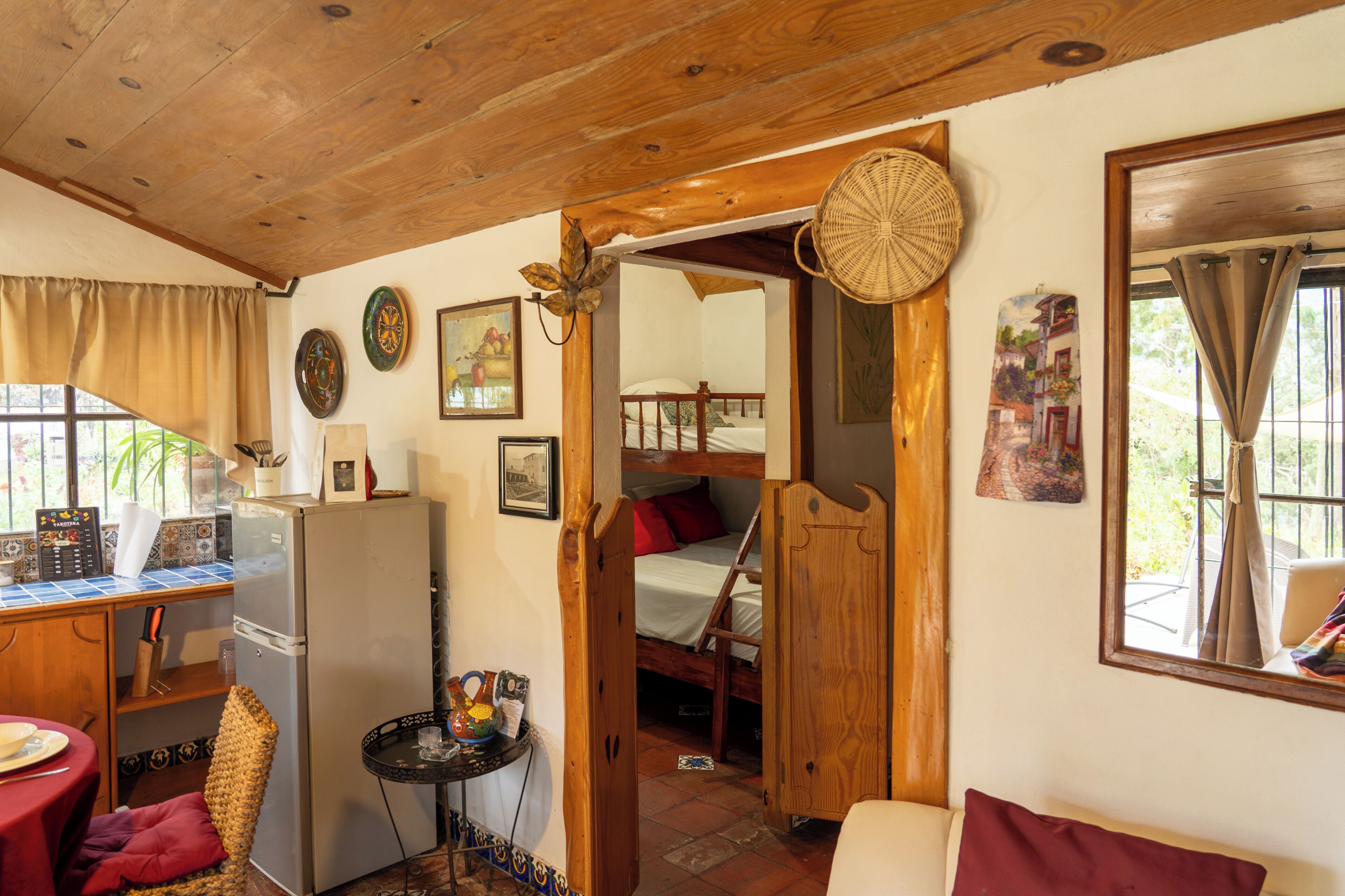 Quarto compacto com cama, uma mesinha, geladeira e janela com cortinas.