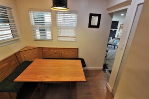 Uma área de refeições com mesa de madeira e banco, duas janelas com persianas e vista para outro cômodo com piso estampado e uma cadeira.