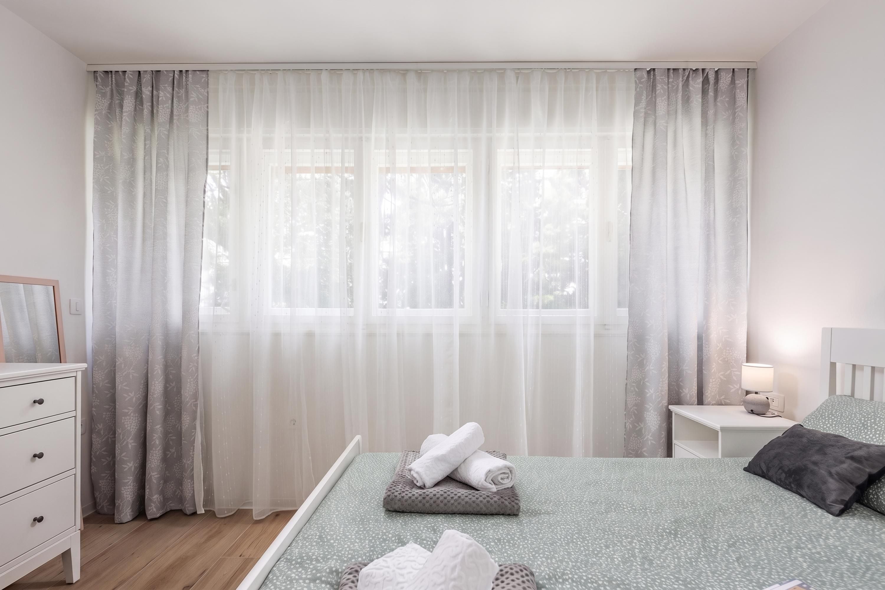 Quarto com cama, mesa de cabeceira, cômoda e janela com cortinas leves.