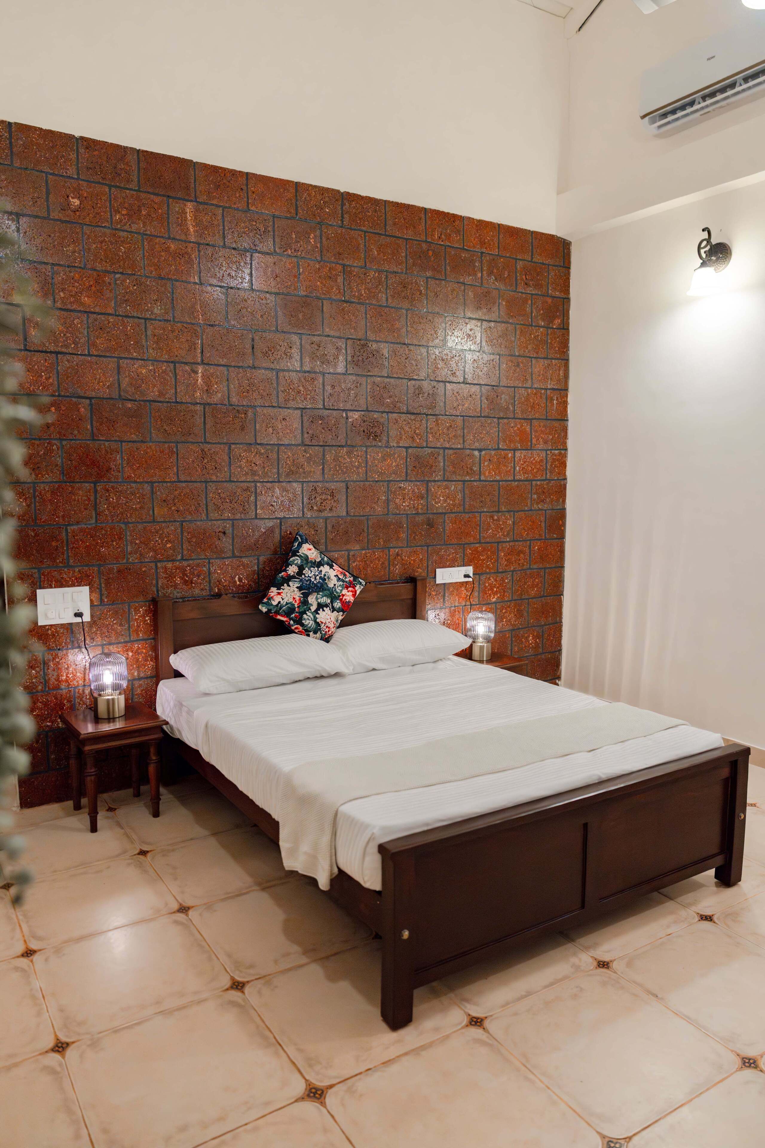 Quarto com cabeceira em tijolos, cama com roupa de cama branca, duas luminárias de cabeceira e mesa de cabeceira com abajur.