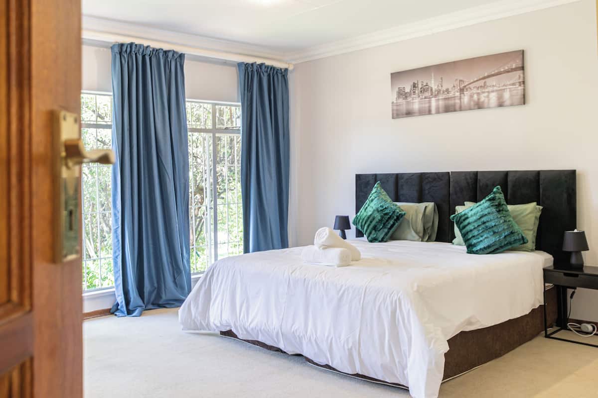 Quarto com uma cama grande, cortinas azuis, pintura de paisagem urbana e mesa de cabeceira com abajur.