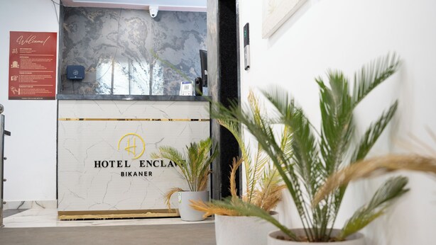 Um balcão de recepção com uma placa indicando "HOTEL ENCLAVE BIKANIER", um código QR e uma placa de boas-vindas.