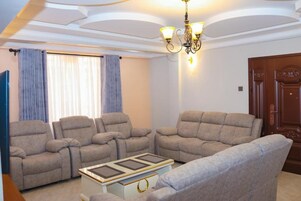 Uma sala de estar com quatro sofás bege dispostos em volta de uma mesa de centro, um candelabro e uma porta de madeira.