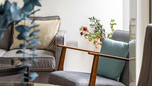 Uma sala de estar moderna com um sofá cinzento, uma mesa de centro de madeira e uma pequena mesa lateral com uma planta em vaso.