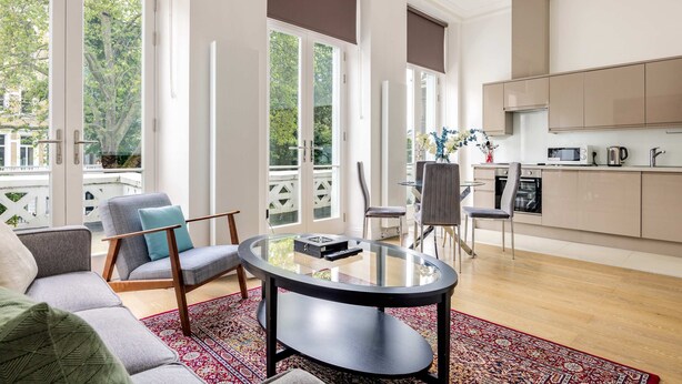 Uma sala moderna com uma mesa de centro de vidro, sofá cinza e um tapete estampado em vermelho. Uma cozinha com armários bege e uma área de jantar com mesa e cadeiras também estão visíveis.