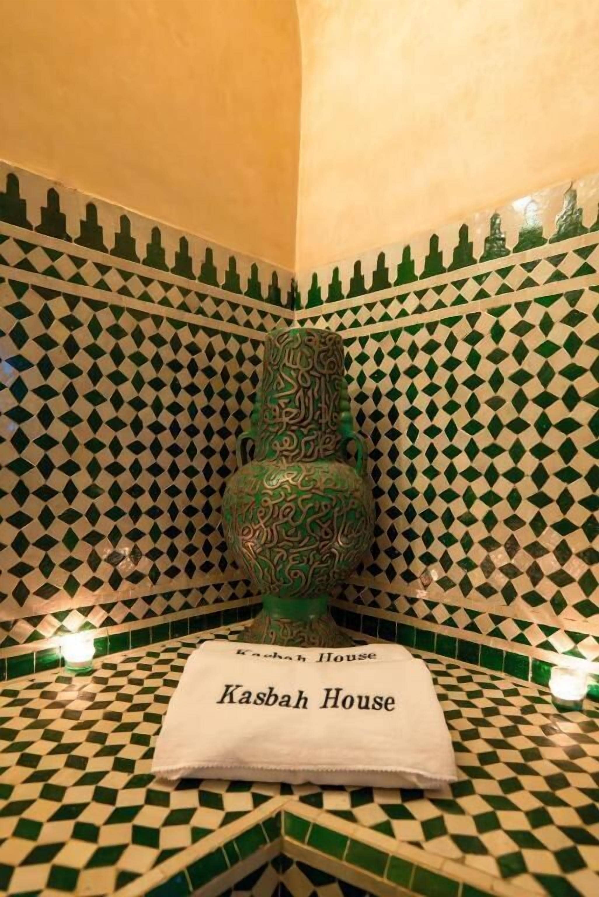 Um quarto com parede de azulejos verde e branco estampados, um vaso decorativo verde e uma toalha no chão com a inscrição "Kasbah House".