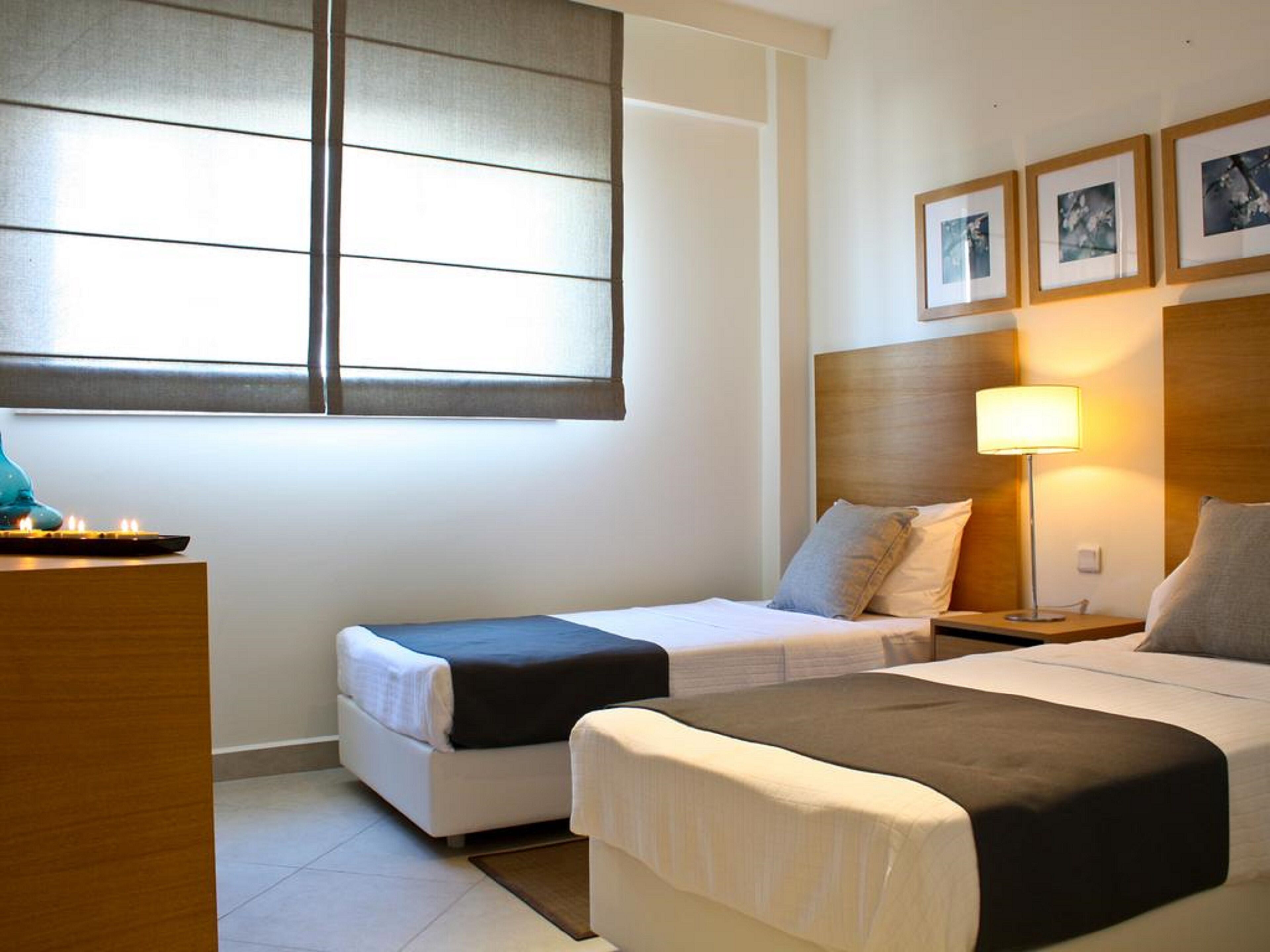 Quarto de hotel com duas camas, mesa de cabeceira com abajur, janela com persianas e quadros nas paredes.