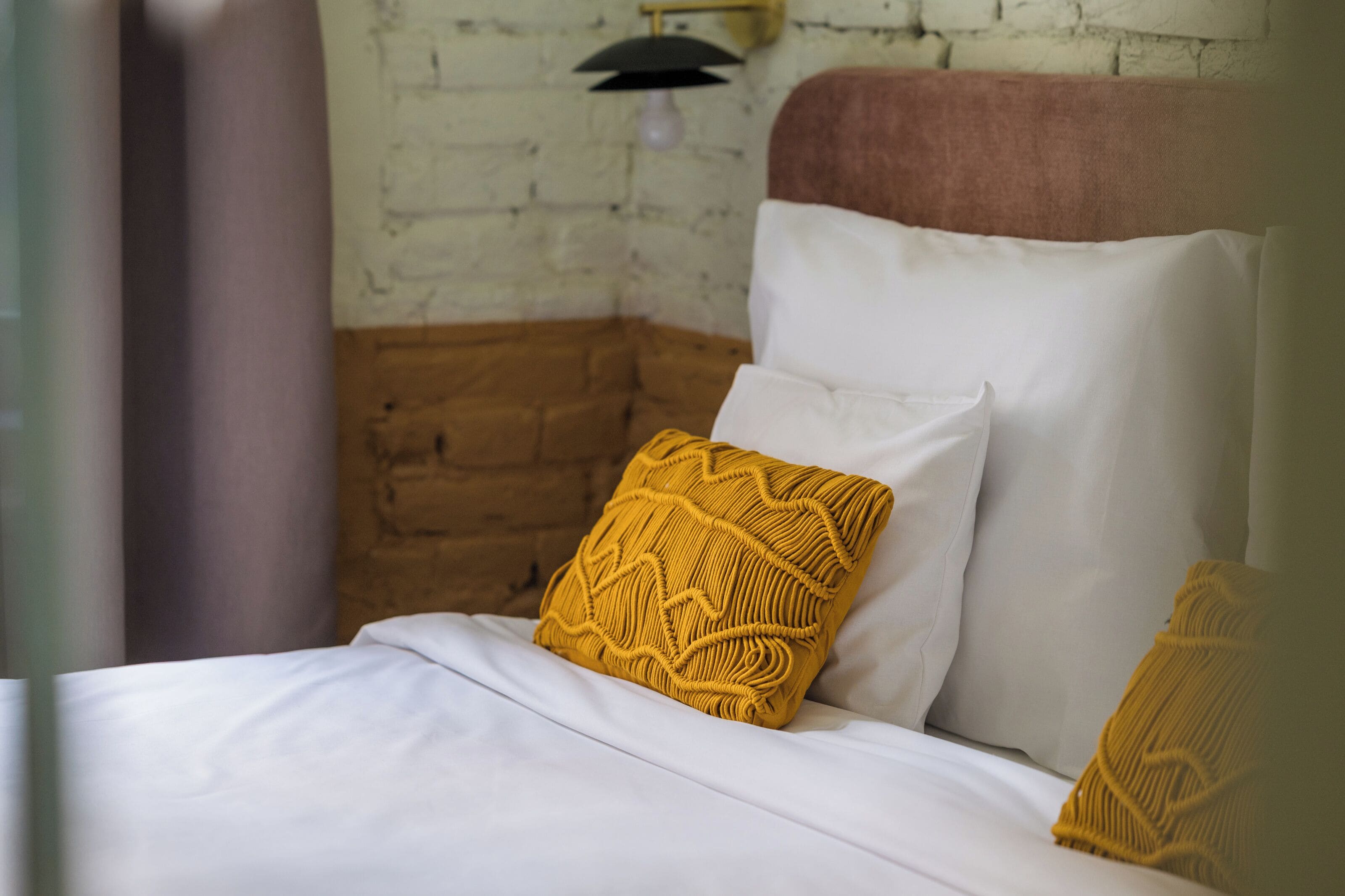 Uma cama com roupa de cama branca e duas almofadas amarelas, uma parede de tijolos e uma luminária ao fundo.