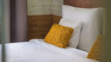 Uma cama com roupa de cama branca e duas almofadas amarelas, uma parede de tijolos e uma luminária ao fundo.