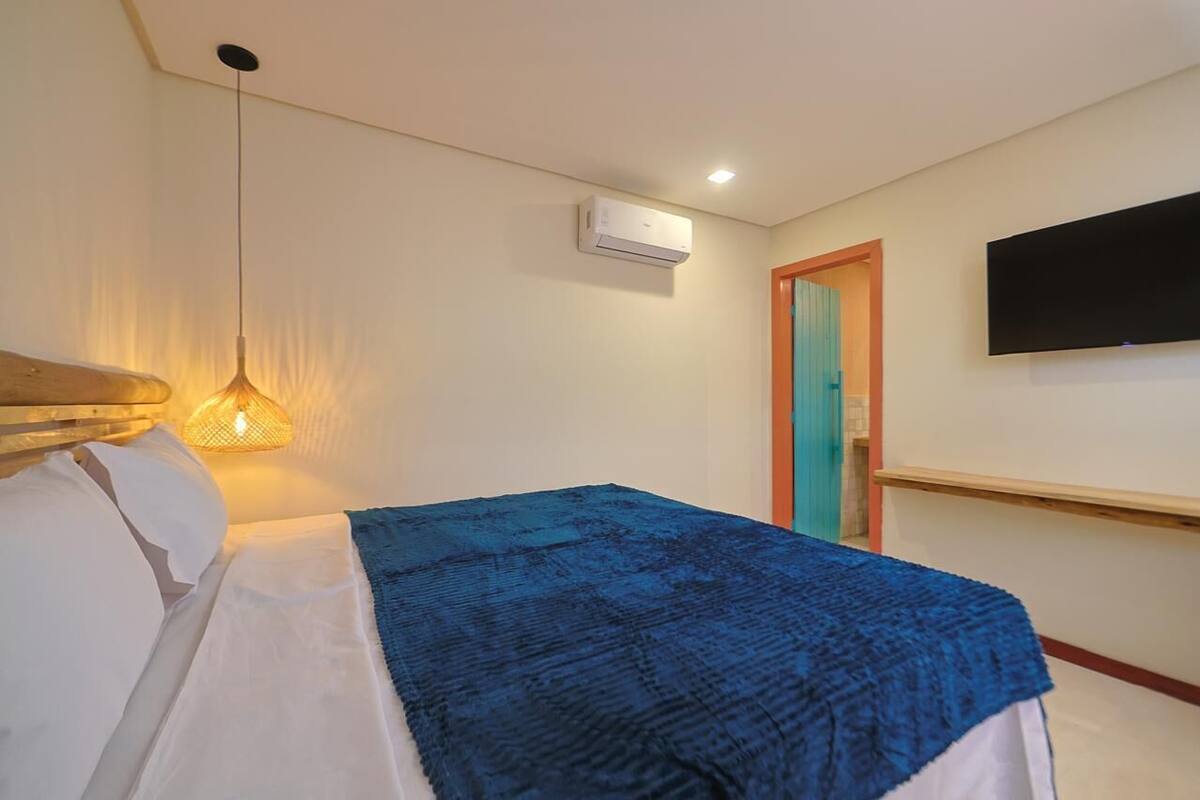 Quarto de hotel com uma cama, luminária pendente, ar condicionado, televisão fixada na parede e uma porta com moldura verde-água.