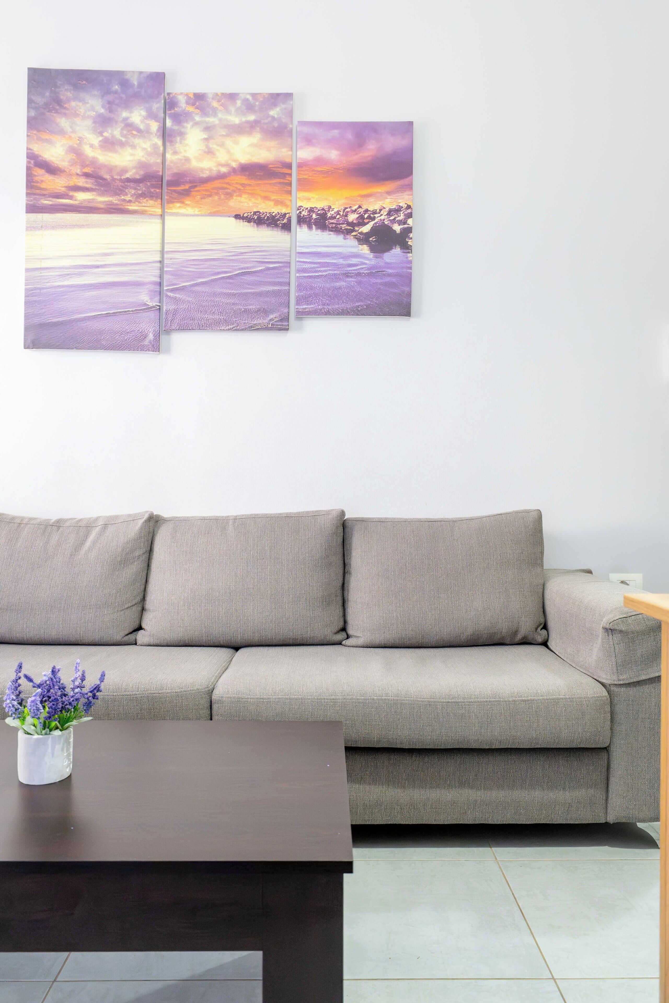 Uma sala de estar com um sofá cinzento, uma mesa de centro e um tríptico de pintura pendurado na parede.