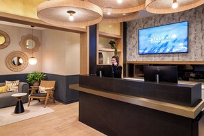 Uma área de recepção com balcão, uma televisão de tela plana grande exibindo 'CATALONA HOTELS & RESORTS' e um espaço de estar com sofá e cadeiras.