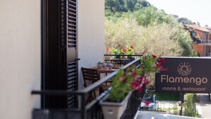 Uma varanda com mesa e cadeiras, um letreiro com 'Flamengo rooms & restaurant' e vista para o verde e edifícios ao fundo.