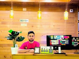 Balcão de recepção com placa indicando área de atendimento, um vaso de planta, um quadro emoldurado e um monitor de computador exibindo informações de redes sociais e um código QR.