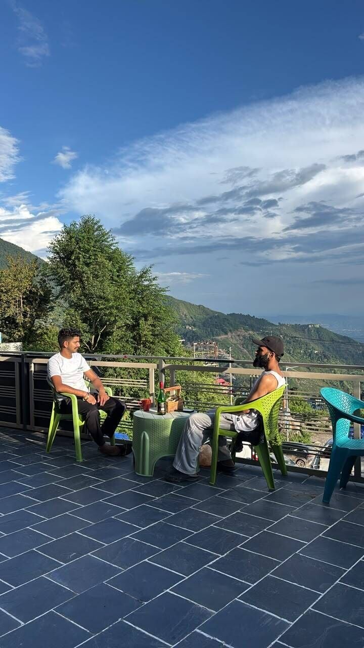 Duas pessoas sentadas em cadeiras ao ar livre, com uma mesa, apreciando a paisagem montanhosa.
