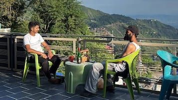 Duas pessoas sentadas em cadeiras ao ar livre, com uma mesa, apreciando a paisagem montanhosa.