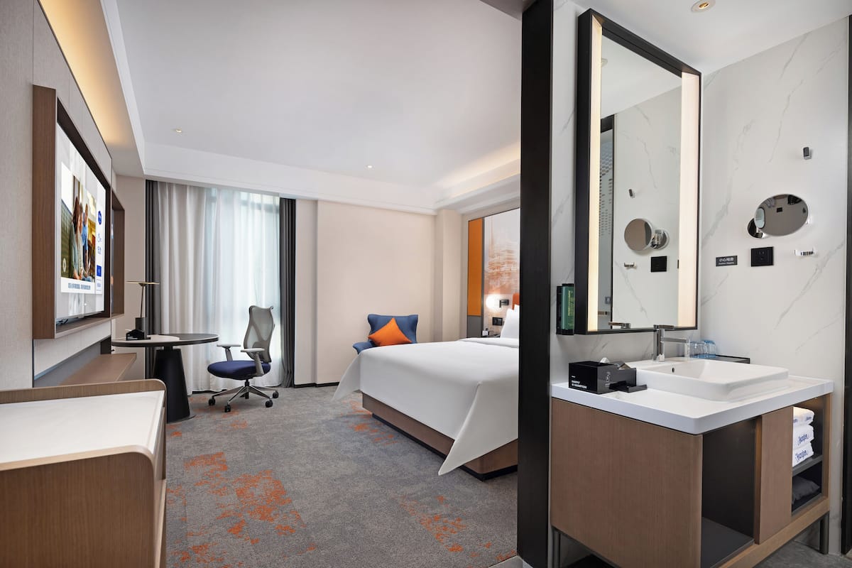 Quarto de hotel moderno com cama, escrivaninha, cadeira, espelho grande, pia e janela com cortinas.