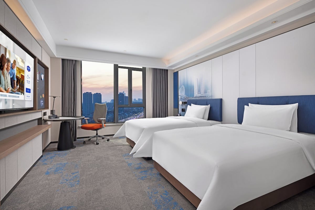 Quarto de hotel com duas camas, uma escrivaninha e uma cadeira. Há uma janela grande com vista para a cidade.