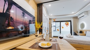 Quarto de hotel moderno com uma grande televisão de tela plana, uma mesa de jantar com um prato de comida e taças de vinho, uma cama bem arrumada e um vaso com flores.