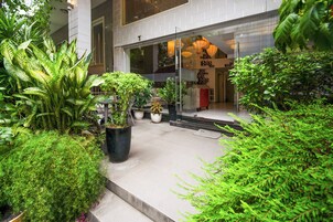 Entrada de hotel com porta de vidro, plantas em vasos e caminho de concreto até a entrada.