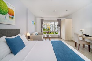 Quarto de hotel com cama, escrivaninha, cadeira, mesa pequena e vista para edifícios pela janela.