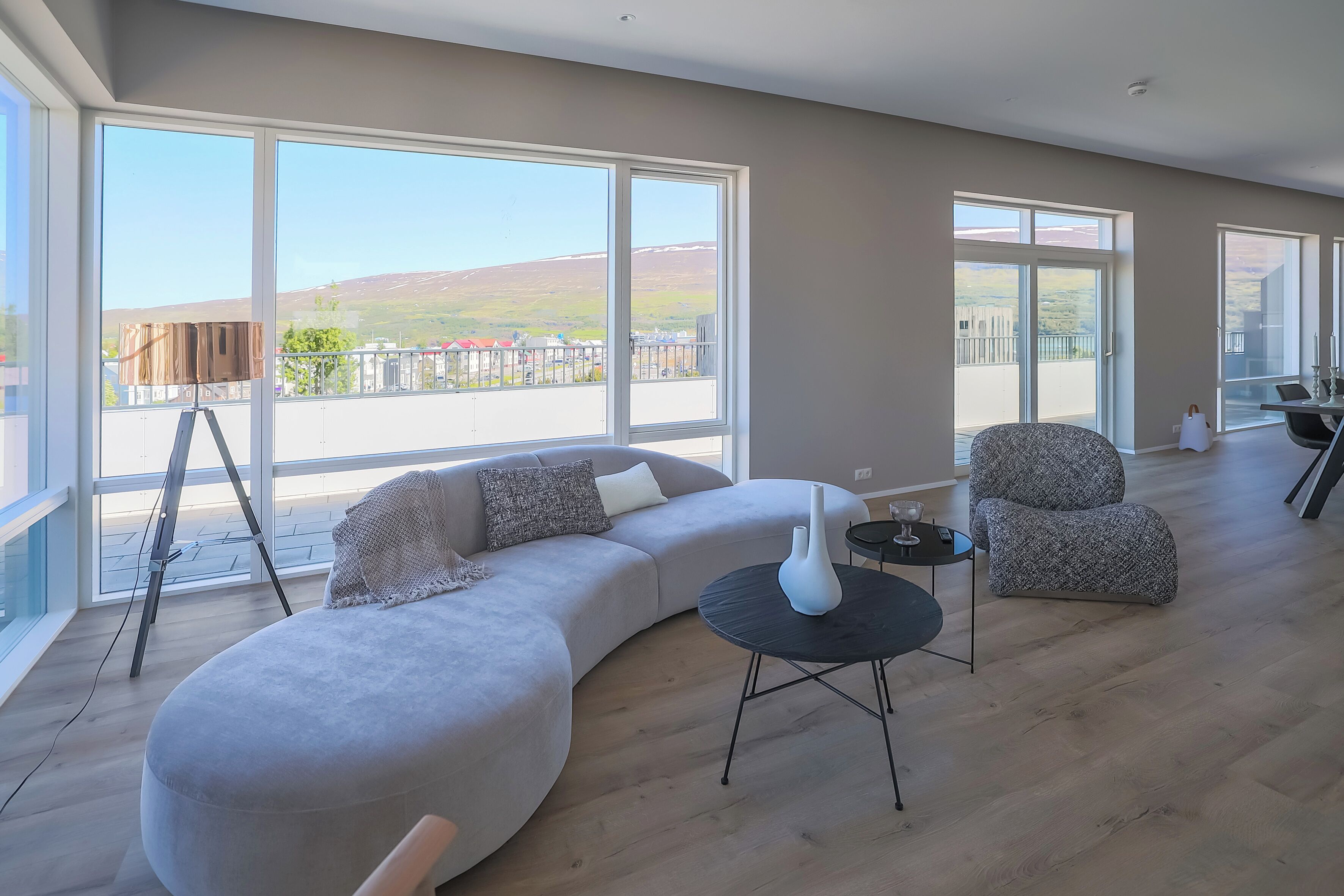 Uma sala de estar espaçosa com um amplo sofá cinza em formato de L, uma mesa de centro redonda preta e uma poltrona estampada. Grandes janelas proporcionam vista para uma varanda e colinas ao longe.