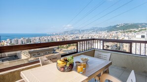 Varanda com mesa posta para dois, com uma fruteira, um recipiente com petiscos e duas taças de bebida. A varanda tem vista para um horizonte urbano com edifícios e uma vista distante do mar.