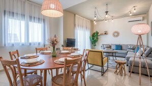 Uma sala de estar moderna com área de jantar, apresentando uma mesa de madeira redonda, cadeiras de vime, um sofá e uma TV.