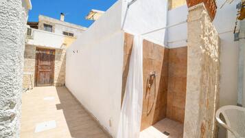 Um corredor exterior estreito com parede branca, porta de madeira e área de duche com parede revestida a azulejos.