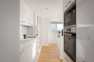 Cozinha moderna com armários brancos, piso de madeira e vista para o mar através de uma janela ampla.
